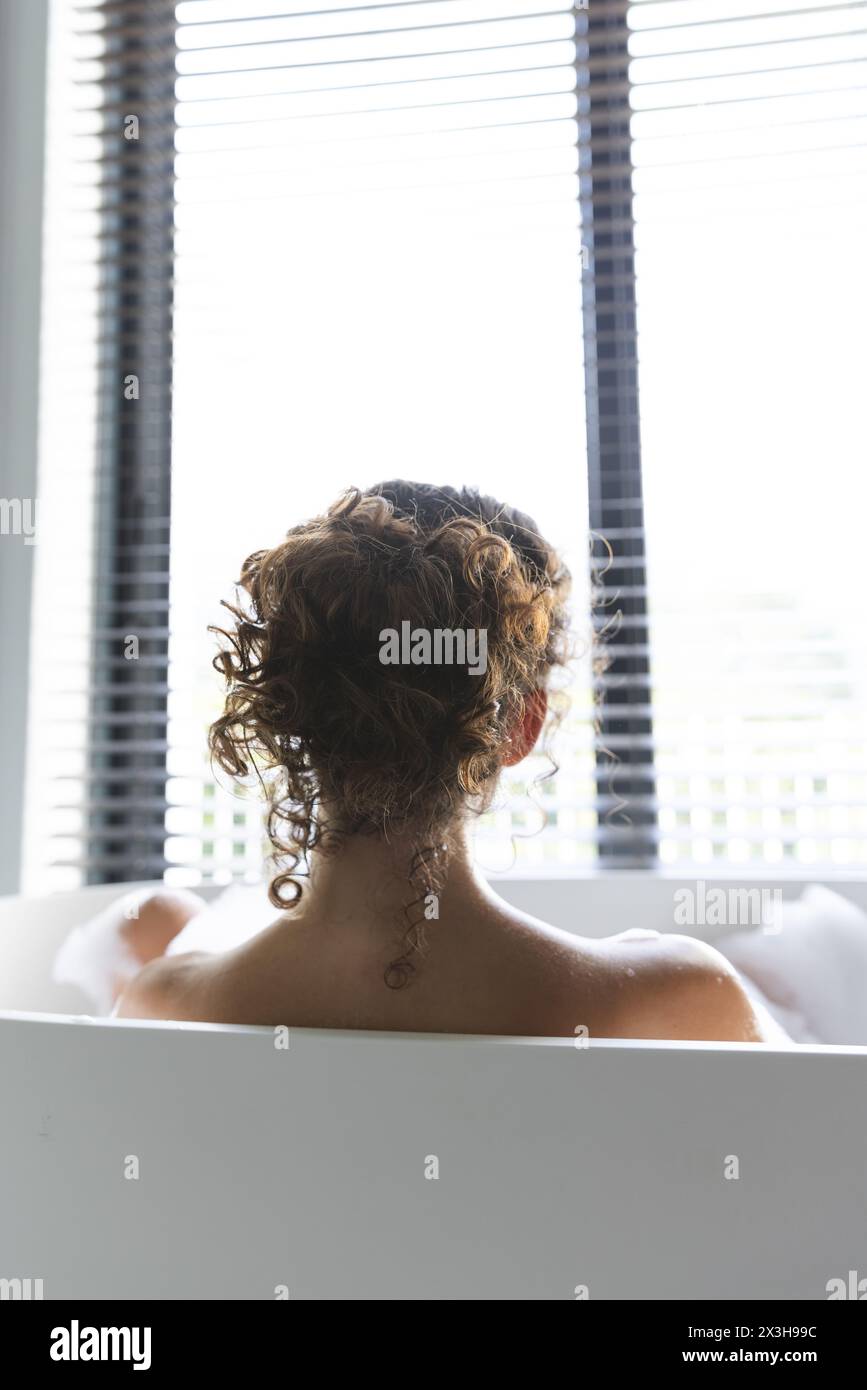 A young Caucasian woman relaxing in bathtub at home, back facing camera ...