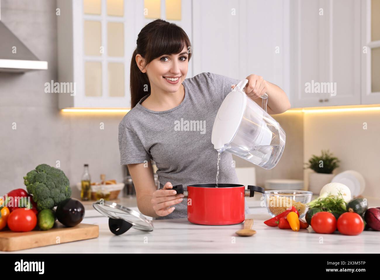 Happy young housewife pouring water in pot at white marble table in ...