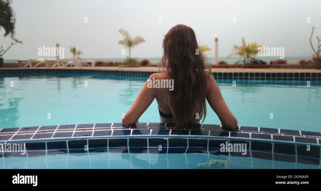 A woman is seated in a swimming pool, facing away from the camera, in a ...