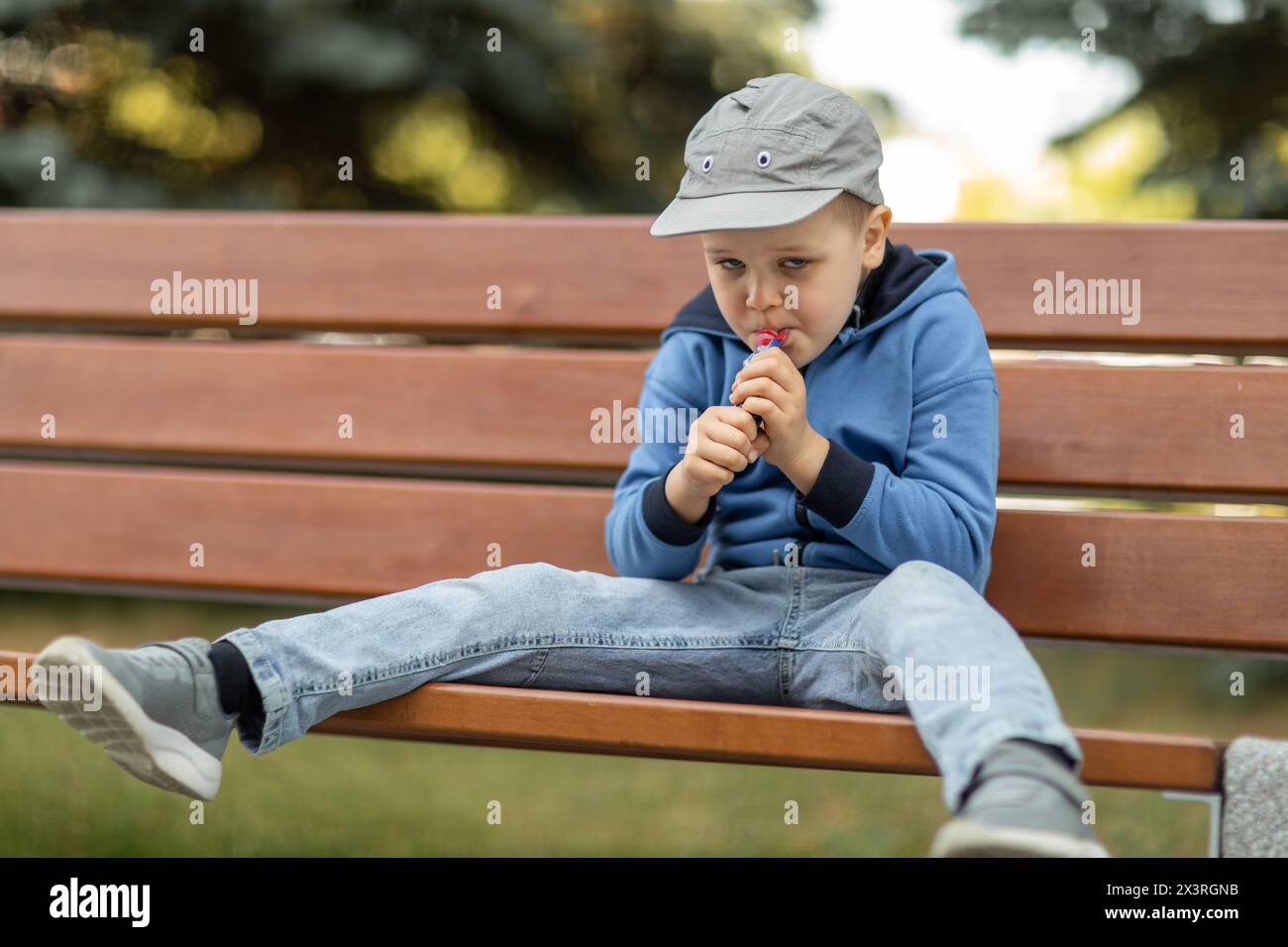 A little boy sitting with legs apart on a brown bench in a city park ...