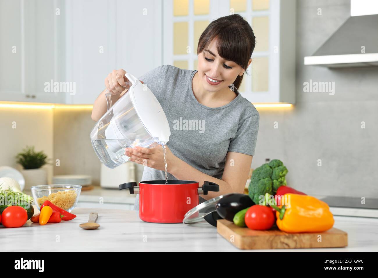 Happy young housewife pouring water in pot at white marble table in ...
