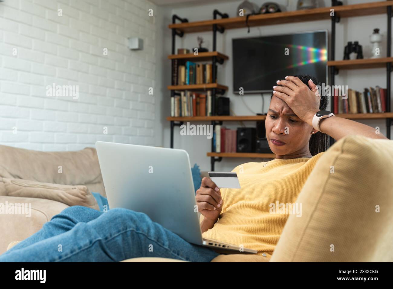 Confused woman sitting on couch holds credit card use laptop looking at ...