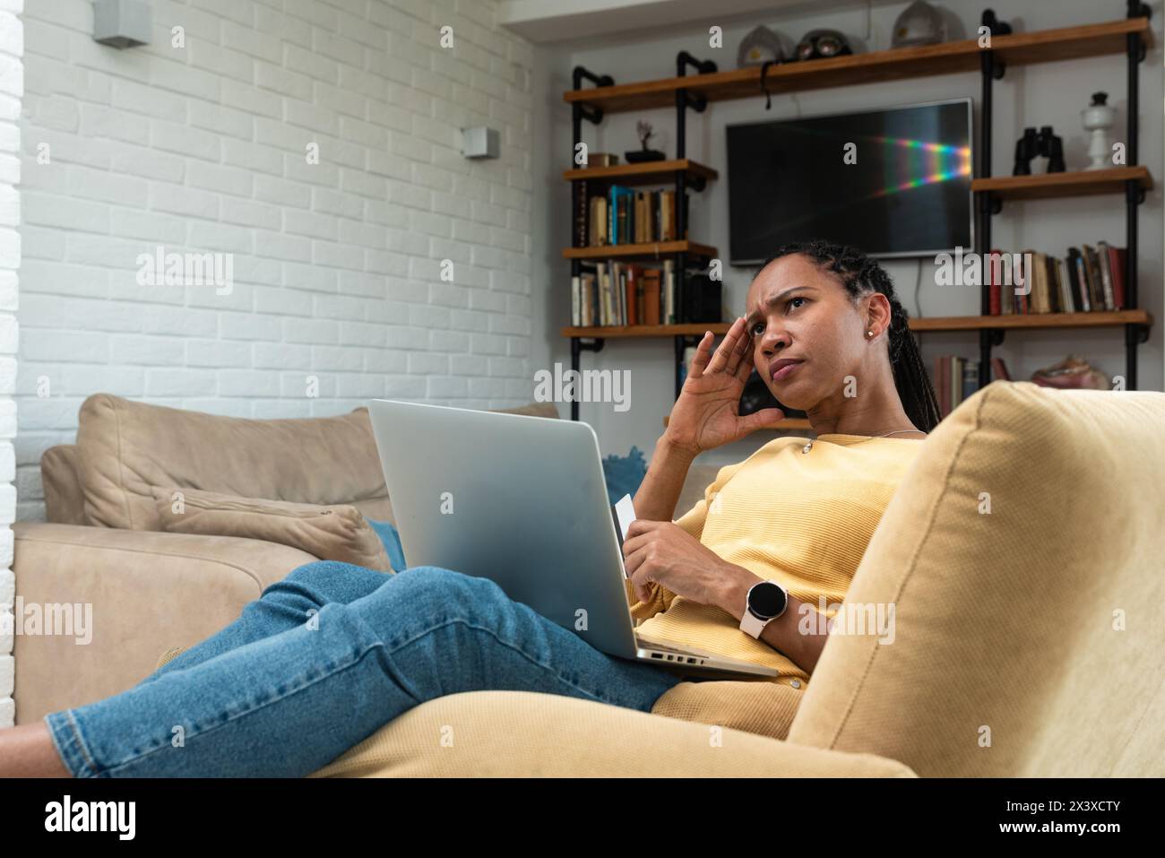 Confused woman sitting on couch holds credit card use laptop looking at ...