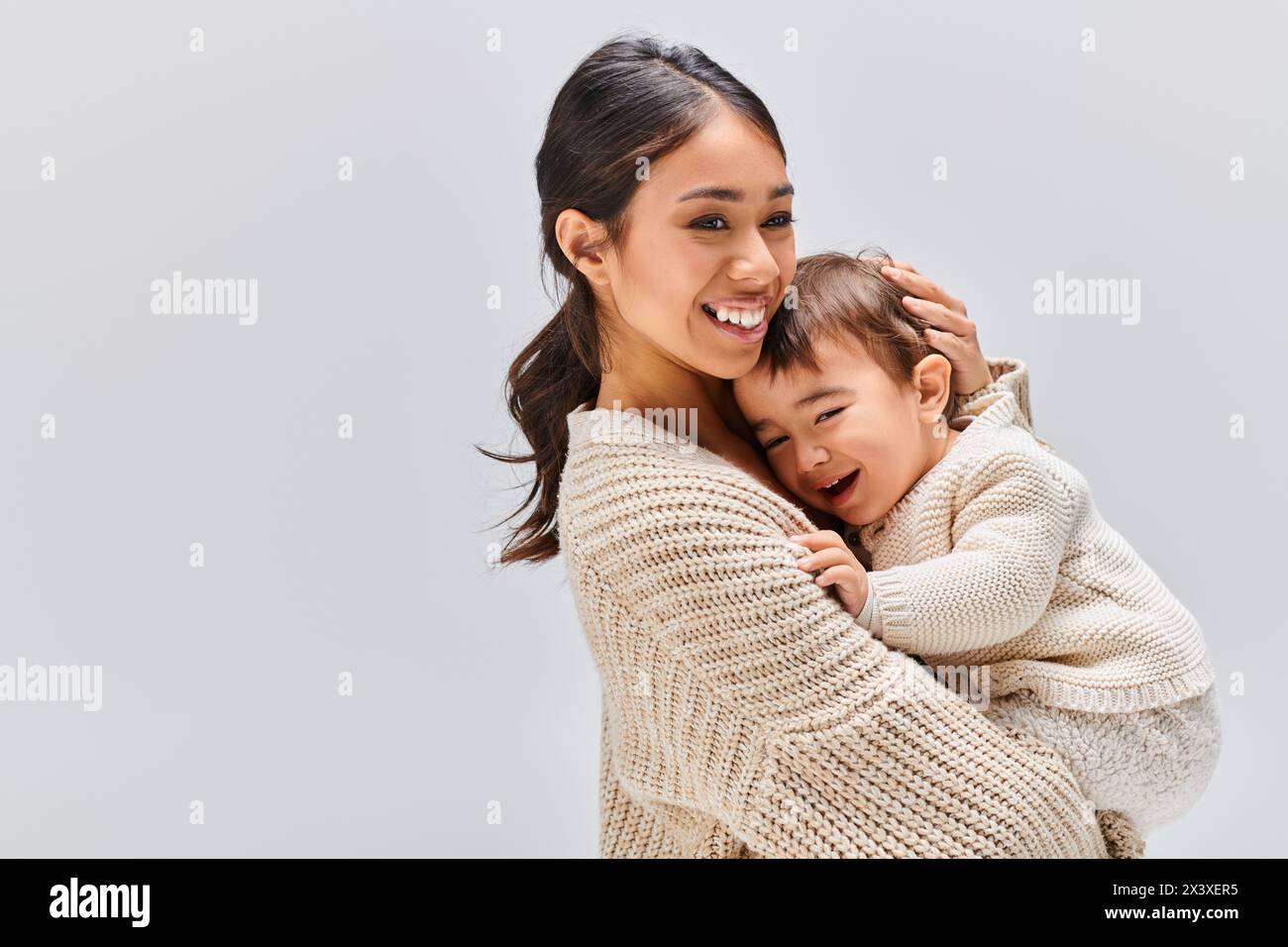 A young Asian mother tenderly embracing her child in a studio setting against a grey background ...