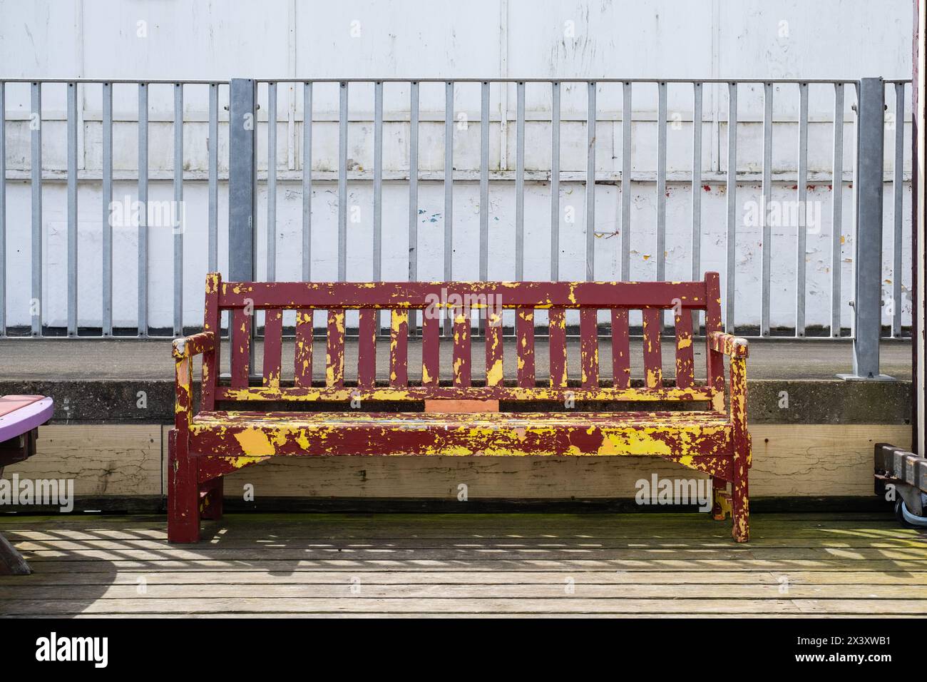 Photograph of a bench with weathered peeling dark red paint revealing yellow paint beneath, on a ...