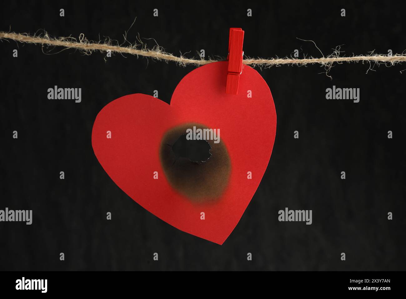 Red paper heart with burnt hole on rope against black background ...