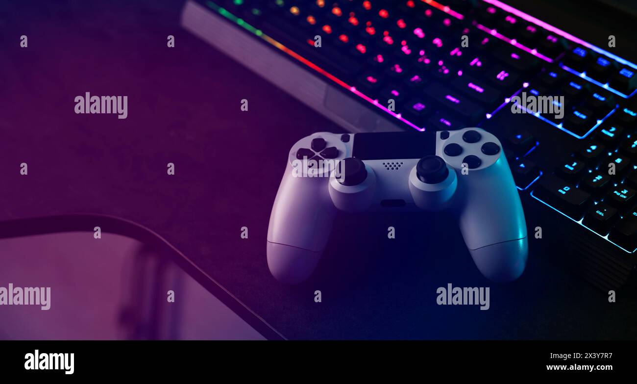 Playing video games. Computer keyboard with RGB lighting and wireless controller on table ...
