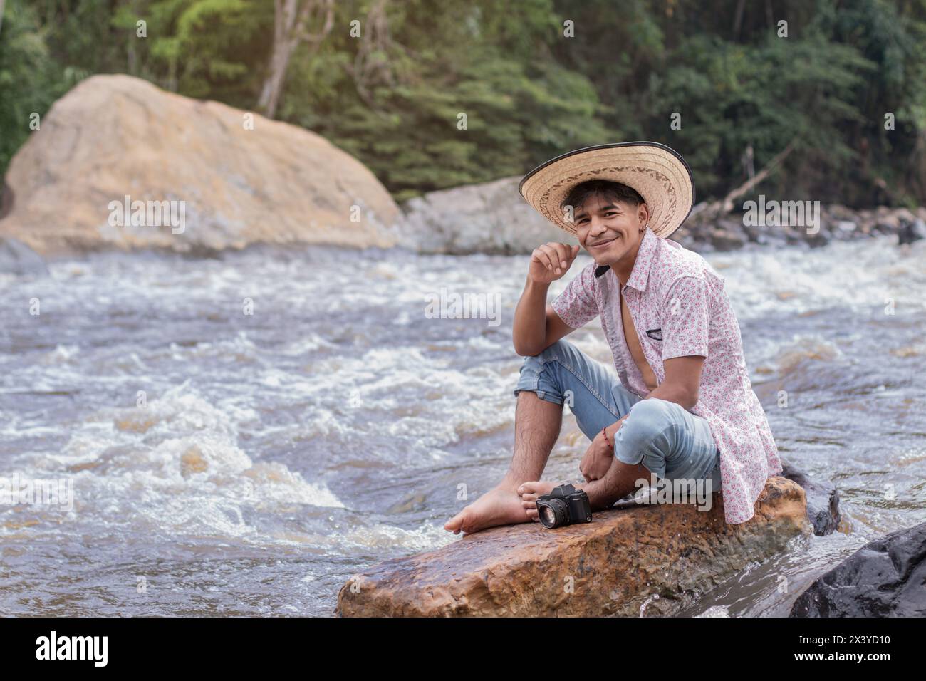 lifestyle. young latin guy wearing a hat sitting on a rock by the river ...