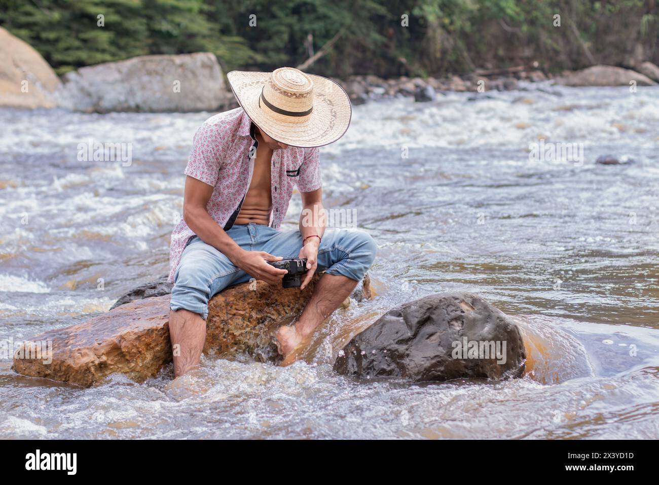 lifestyle. young latin guy sitting on a rock in the river using analog ...