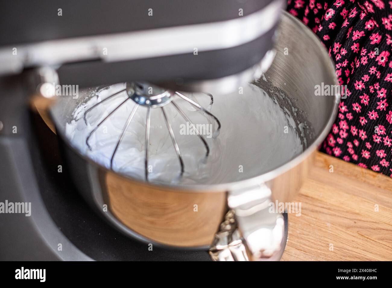 Close up of a stand mixer whipping meringue, with a glimpse of a baker ...