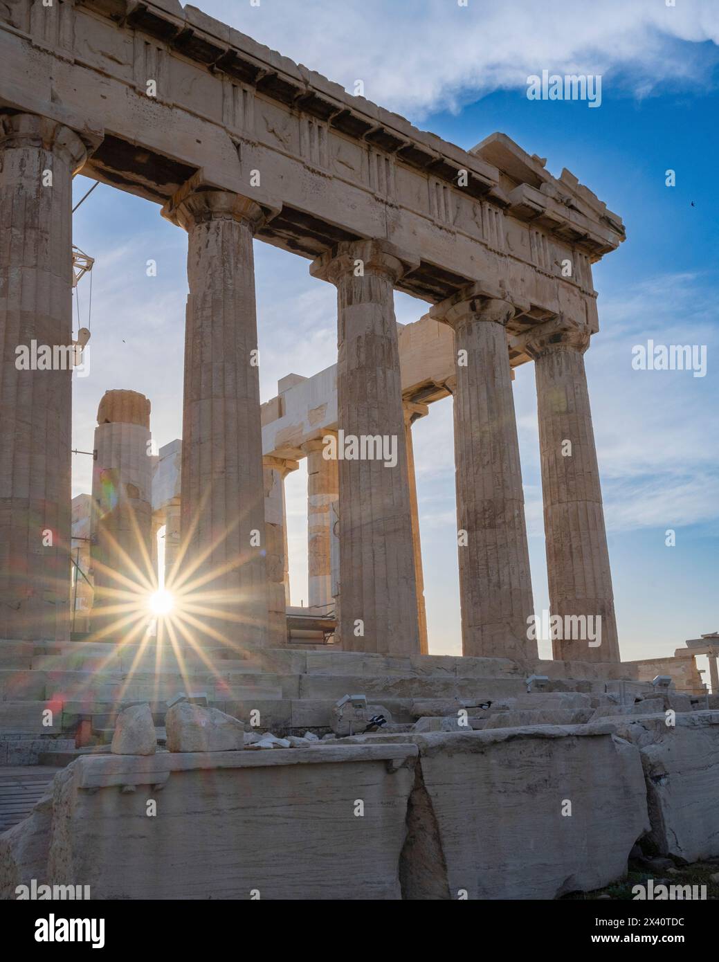 Close-up view of the columns of The Parthenon against a blue, cloudy sky with a sunburst at ...