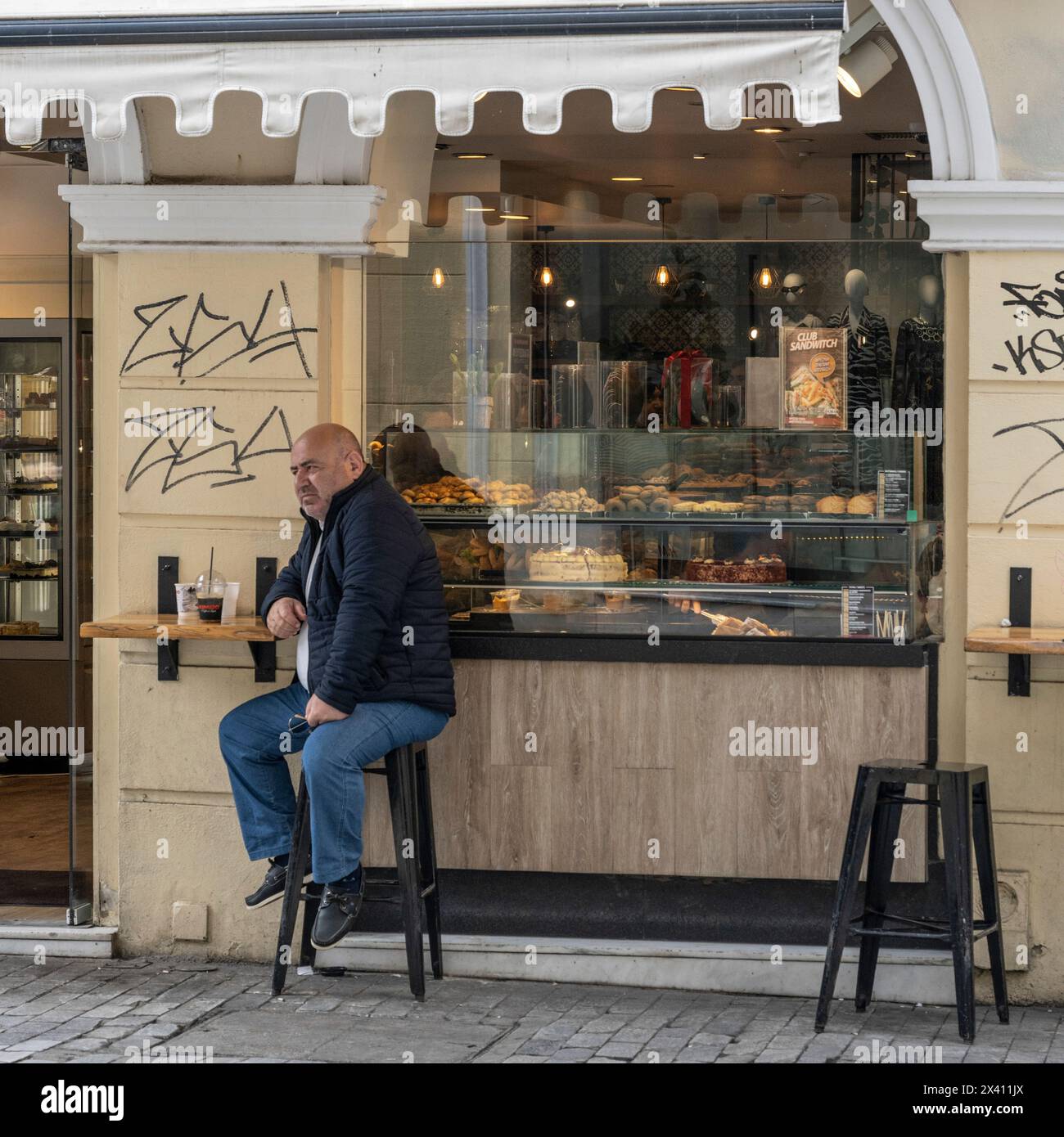 Man sits on a stool under an awning outside a cafe selling pastries ...