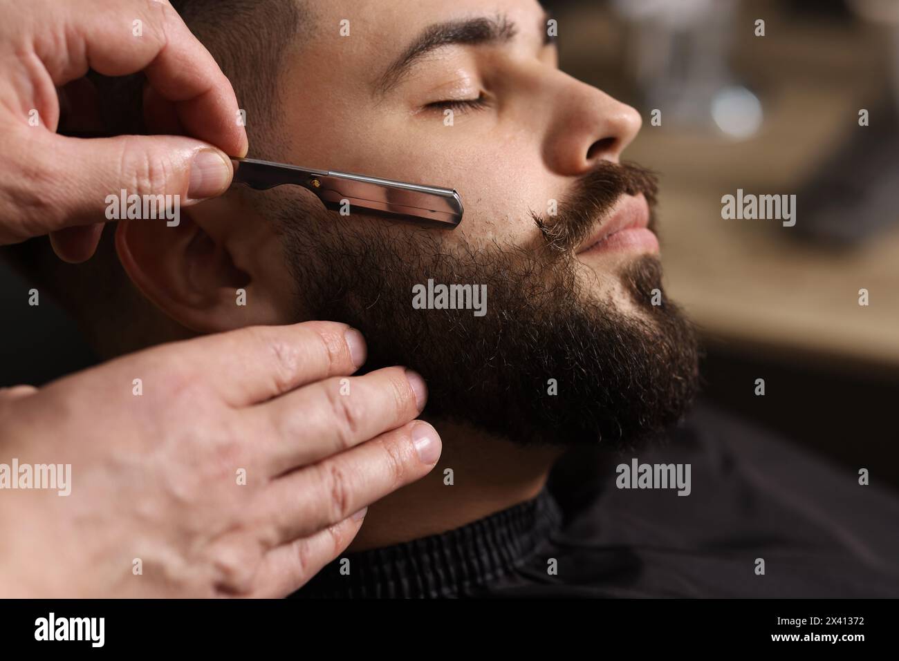 Professional barber shaving client's beard with blade in barbershop ...