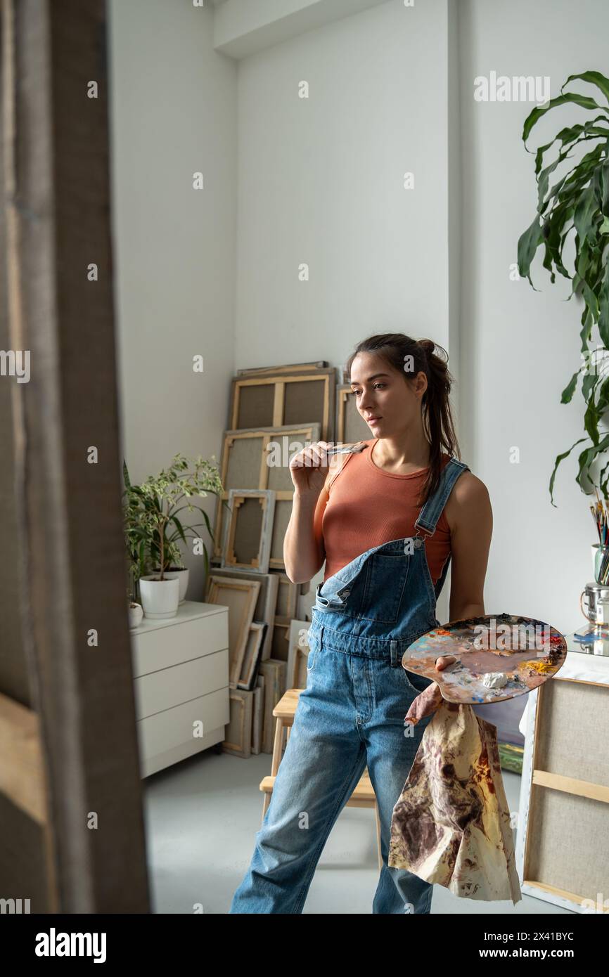 Thoughtful female painter artist standing in front easel with colour ...