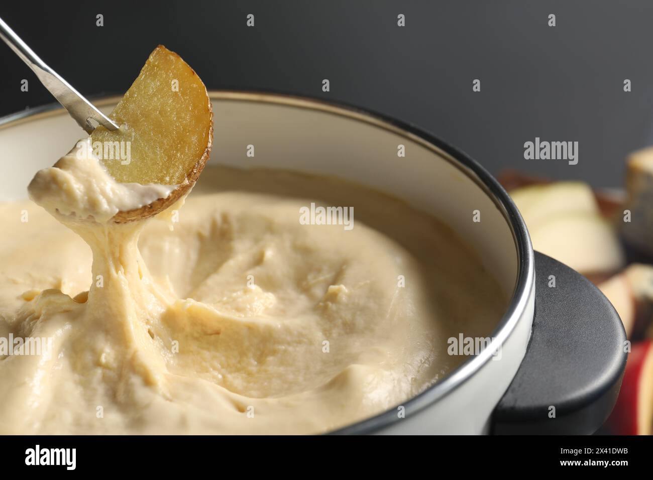 Dipping piece of potato into fondue pot with melted cheese on grey ...