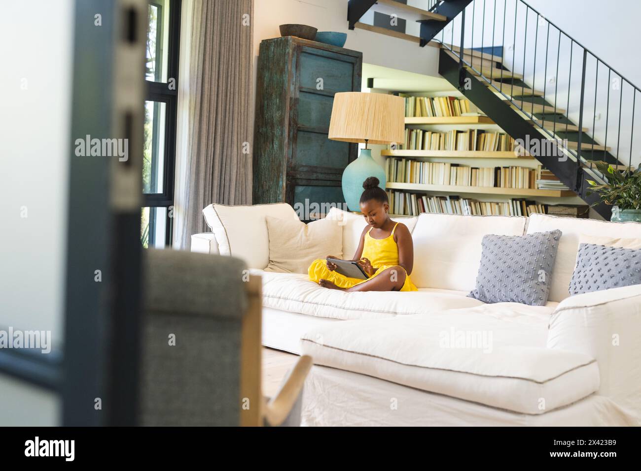 African American girl in yellow dress, reading at home on white sofa ...