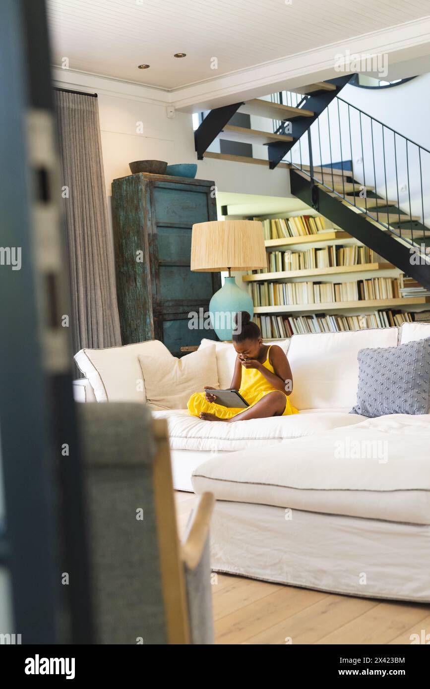African American girl in yellow dress, reading at home on white couch ...