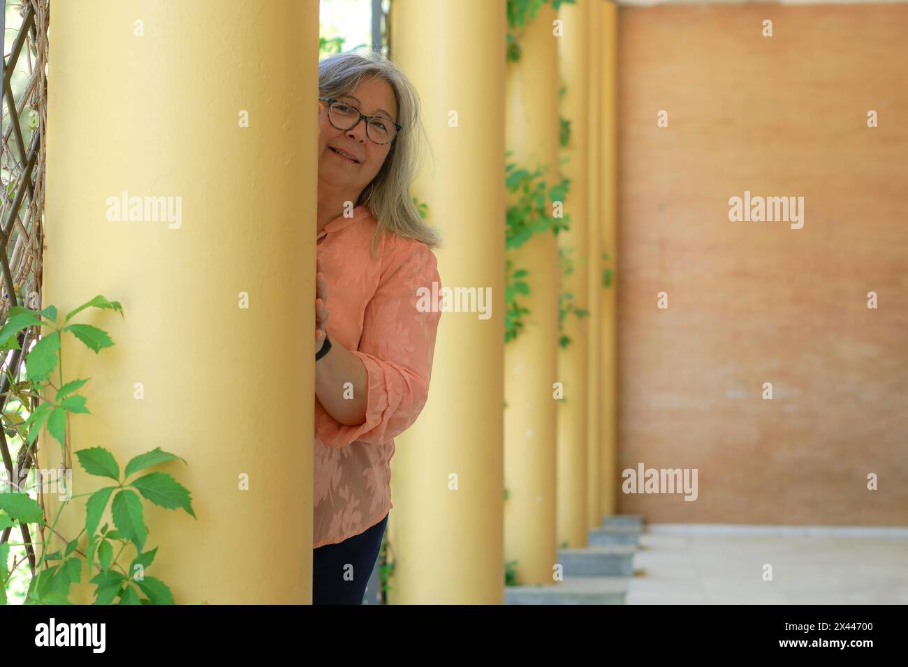 Older woman with white hair and glasses, smiling from behind a column ...