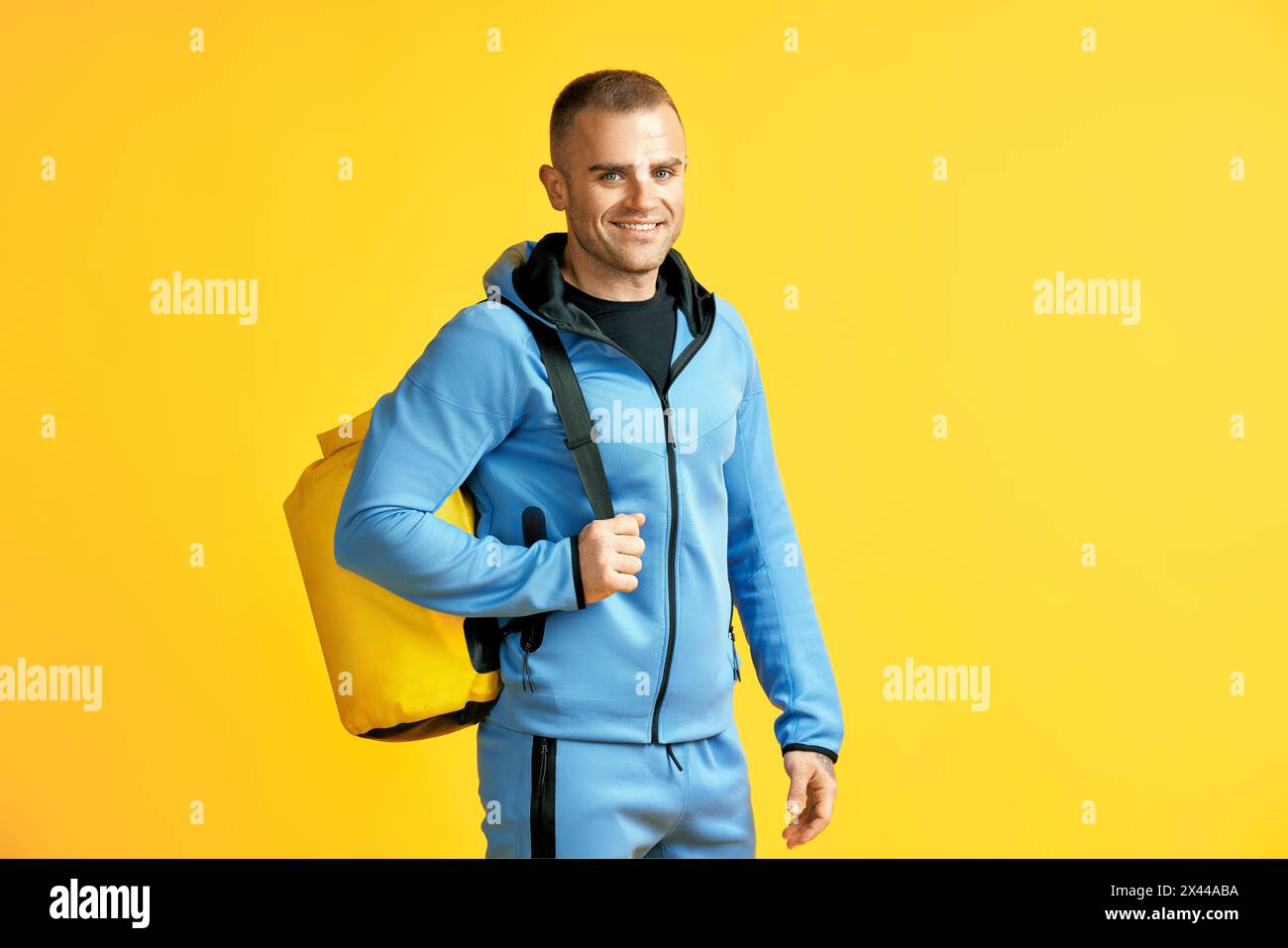 Smiling sporty man in sports suit and back pack over yellow background ...