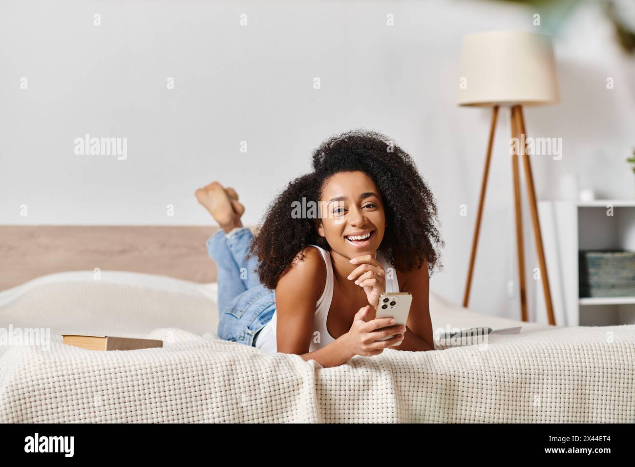 A curly African American woman in a tank top lays on a bed, holding a ...