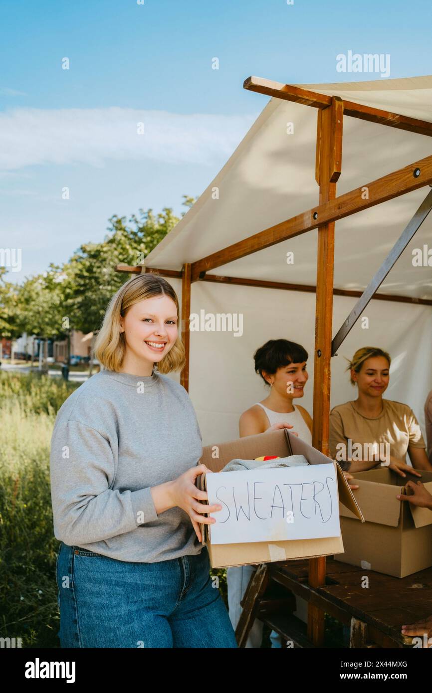 Portrait of happy volunteer holding sweater box during charity drive at ...