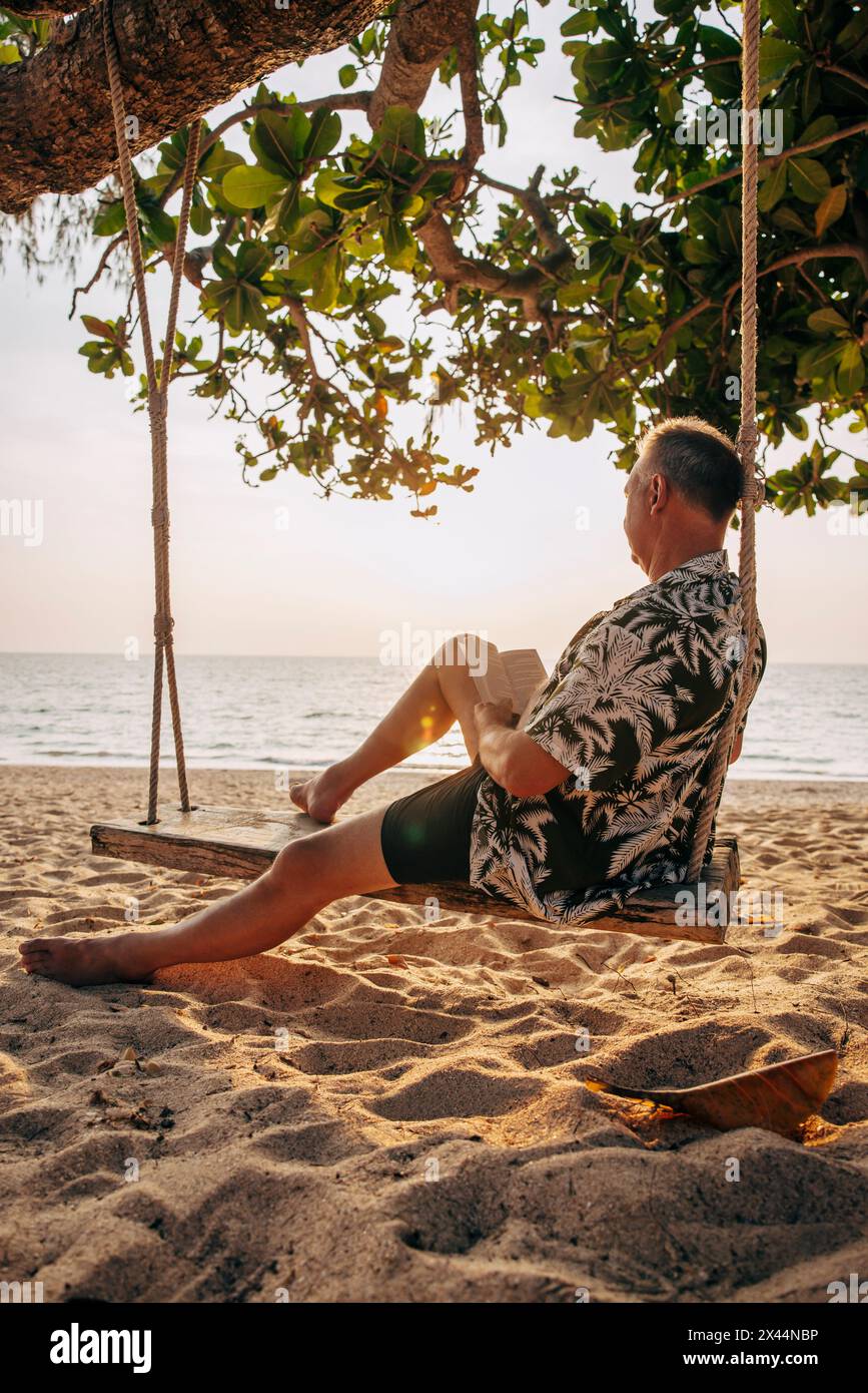 Side view of senior man reading book while sitting on swing at beach ...