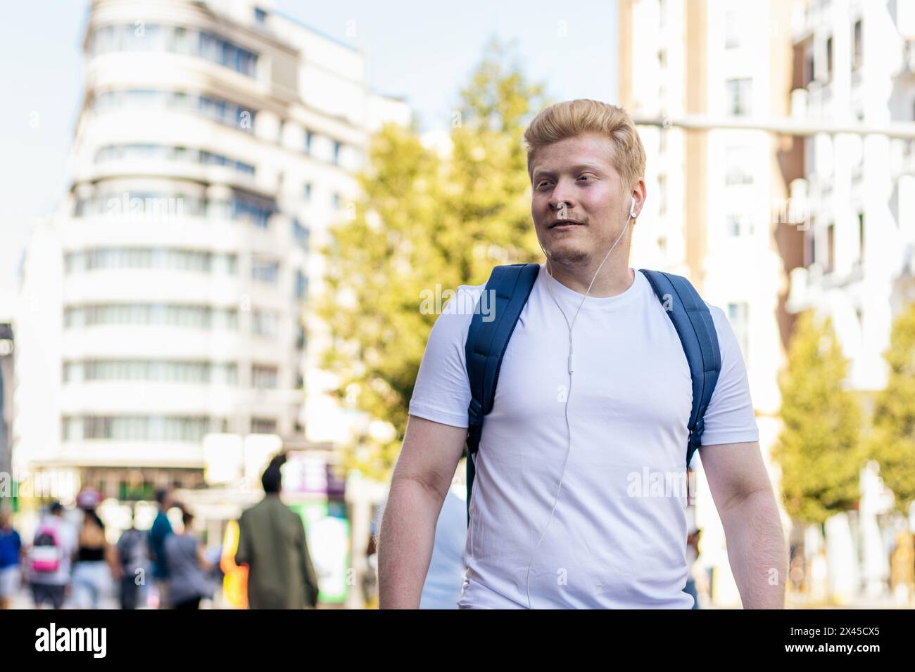 Albino latino man dressed in white clothes and backpack walking in the city street listening to ...