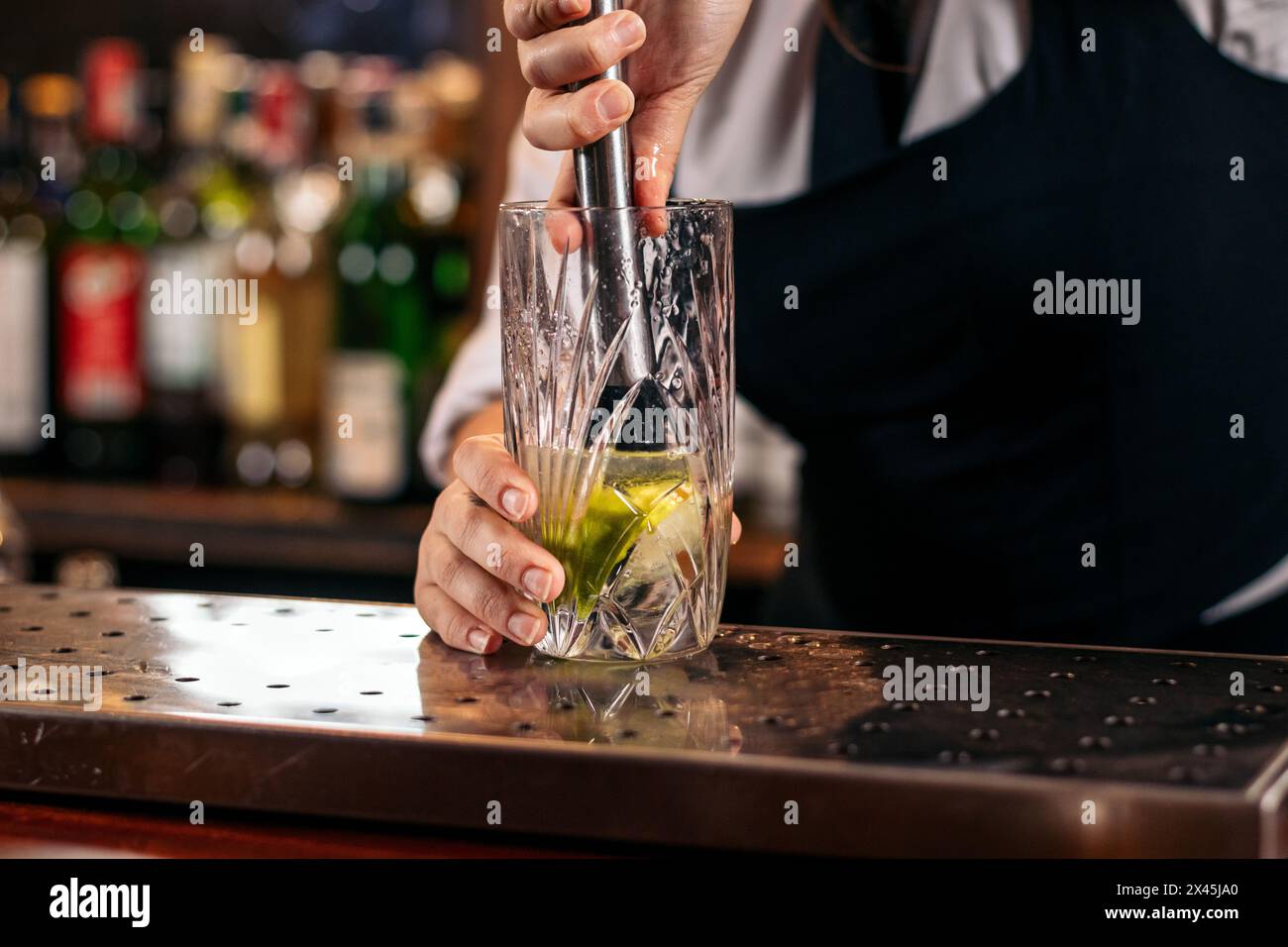 Female bartender preparing a mojito in a tall glass at a cocktail bar. She crushes the lime and ...