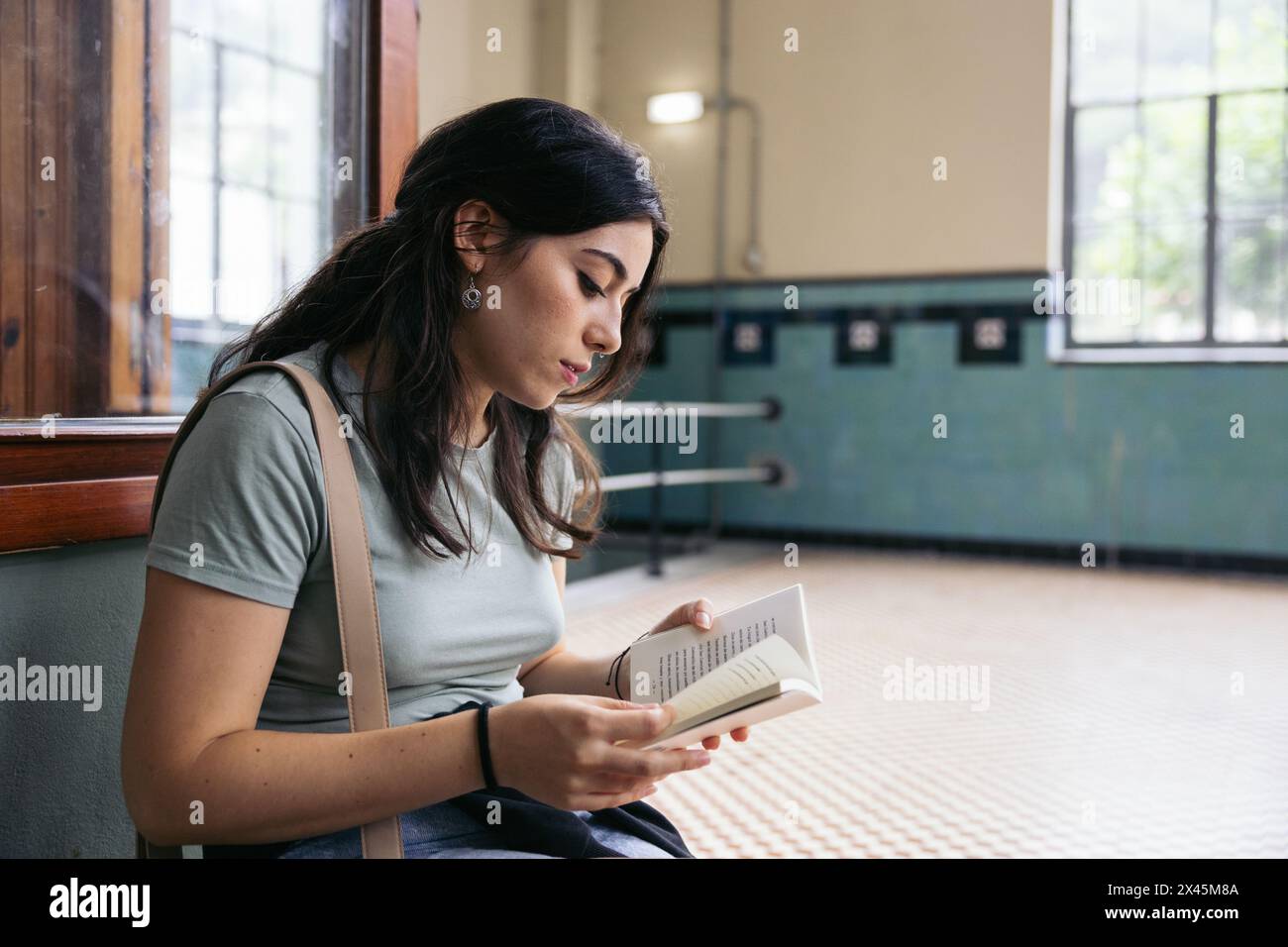A young woman sits on a bench at an old train station, reading a book ...