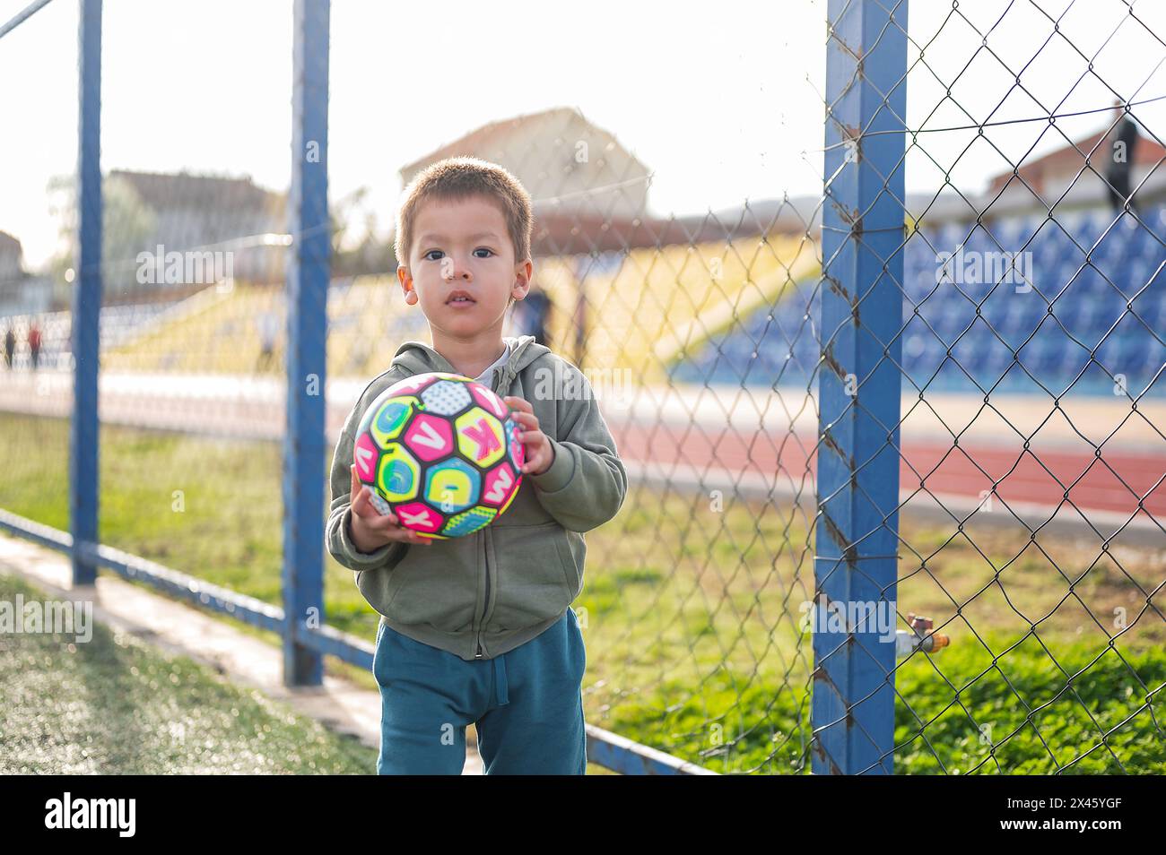 A toddler holding a soccer ball stands by the sports field fence ...