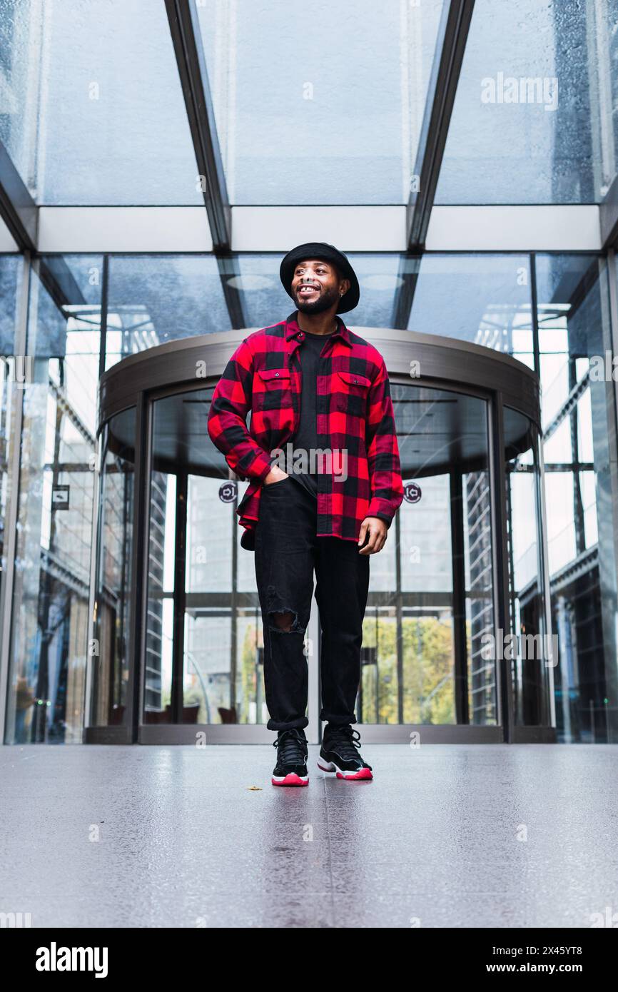 Full body of glad ethnic man in hat walking in hall of modern building ...