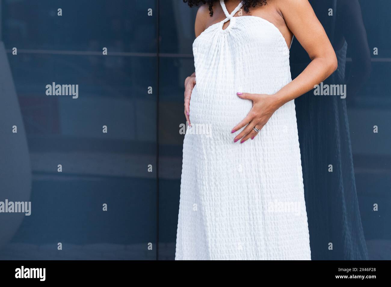 Cropped unrecognizable pregnant African American wearing white dress standing near reflecting ...