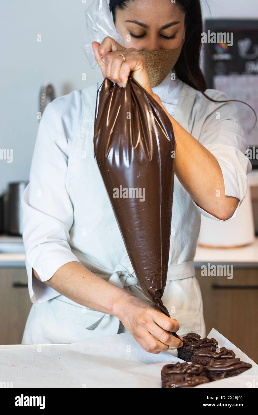 Female baker in mask squeezing chocolate cream from pastry bag on tasty ...