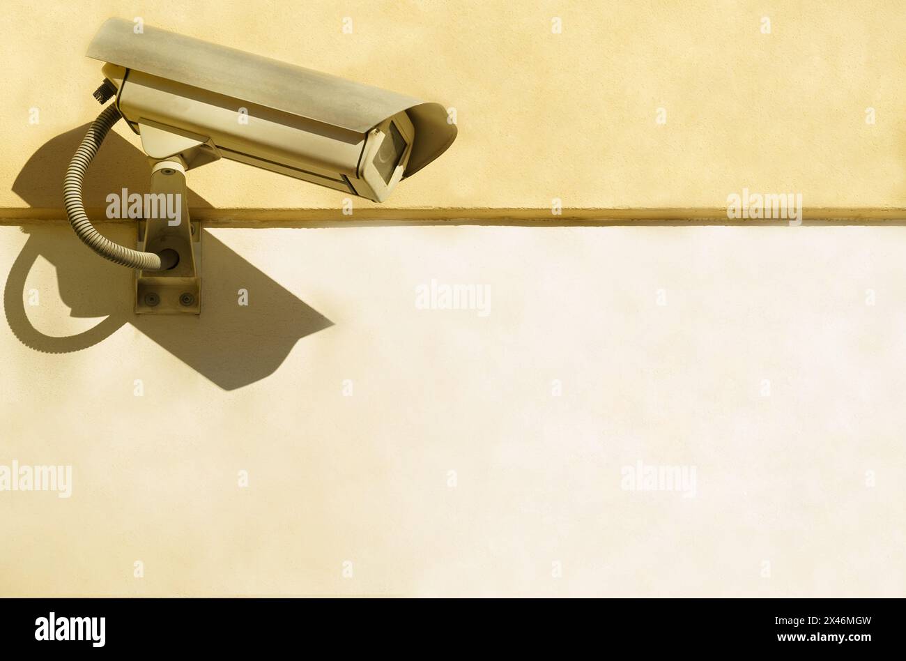 A CCTV camera is affixed to a beige wall, surveilling an open space ...