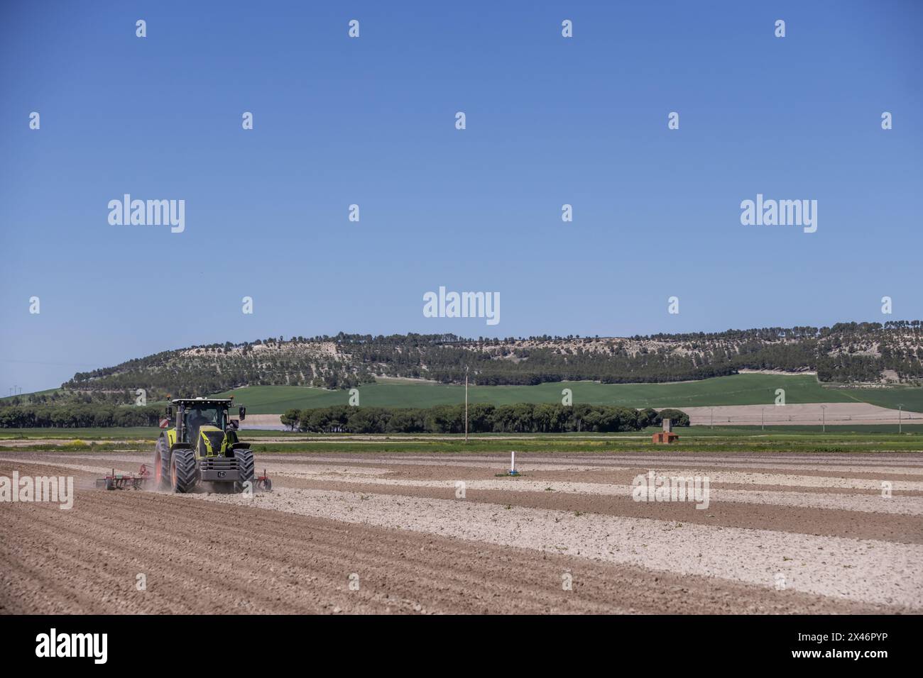 A green tractor pulling a red plow through a crop field with a forested ...