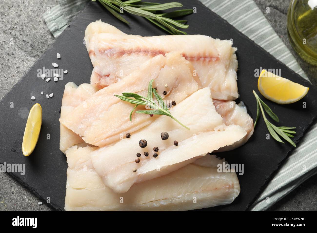 Pieces of raw cod fish, rosemary, peppercorns and lemon on grey table ...