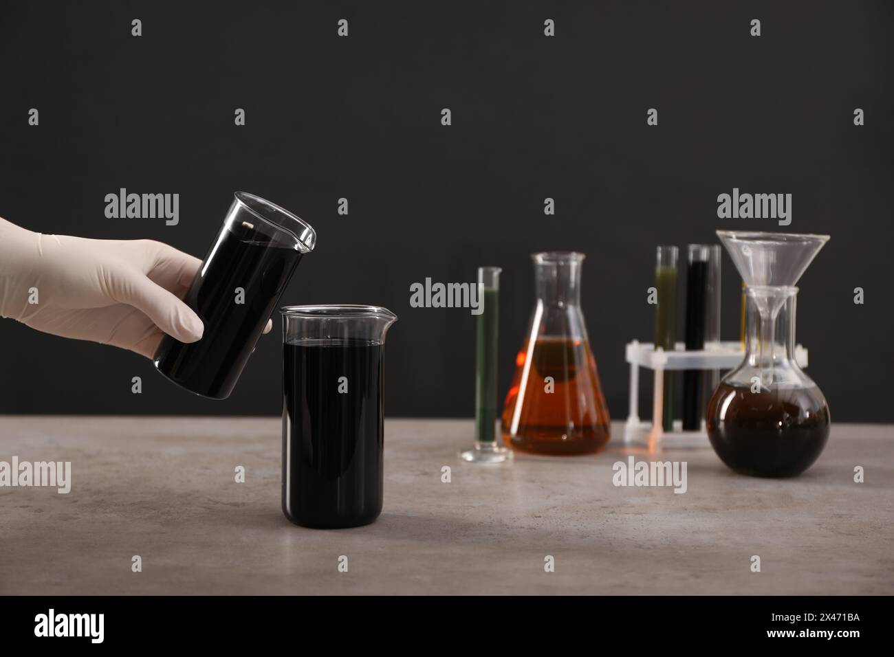 Woman pouring black crude oil into beaker at grey table against dark ...