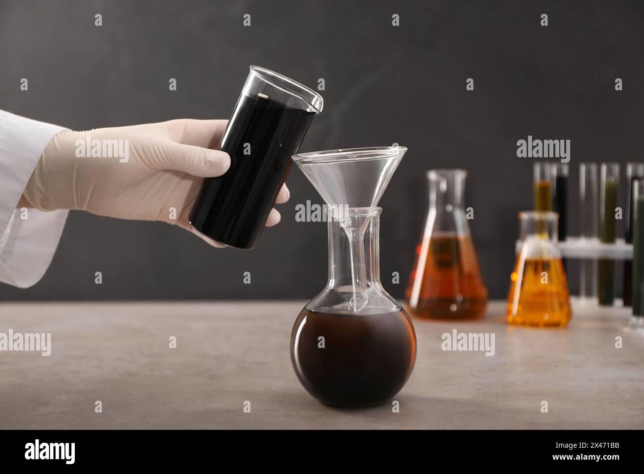 Woman pouring black crude oil from beaker into flask at grey table ...