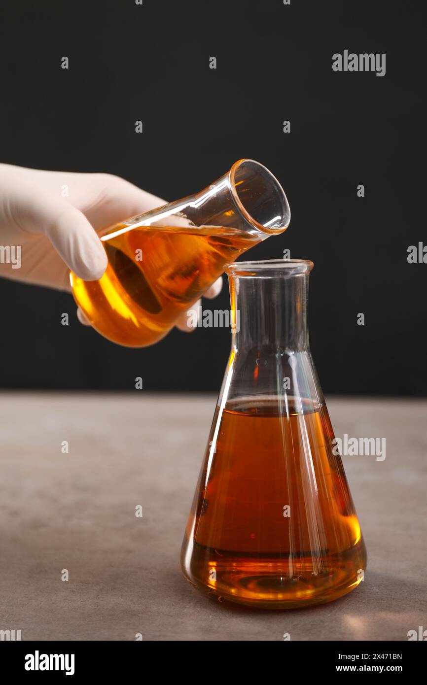 Woman pouring yellow crude oil into flask at grey table against dark ...