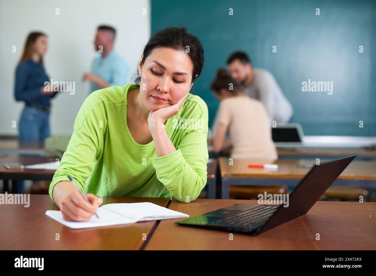 Female student writing lectures in workbooks and laptop in classroom ...