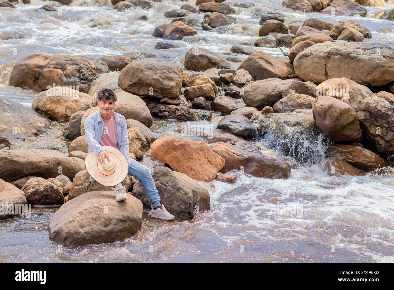 lifestyle. young latin guy sitting on the rocks by the river with hat ...