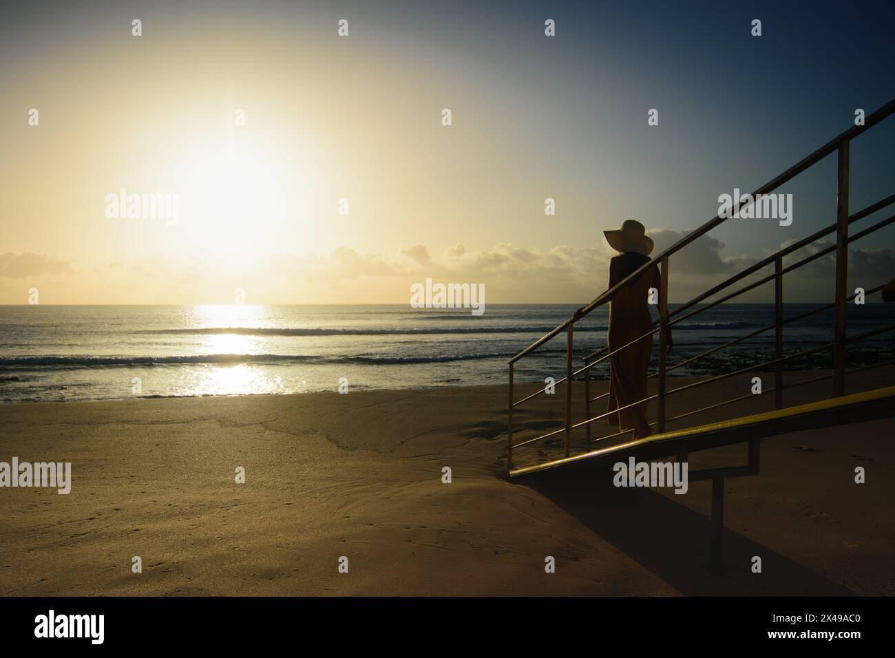 A woman stands on a beach looking out at the ocean. The sun is setting ...
