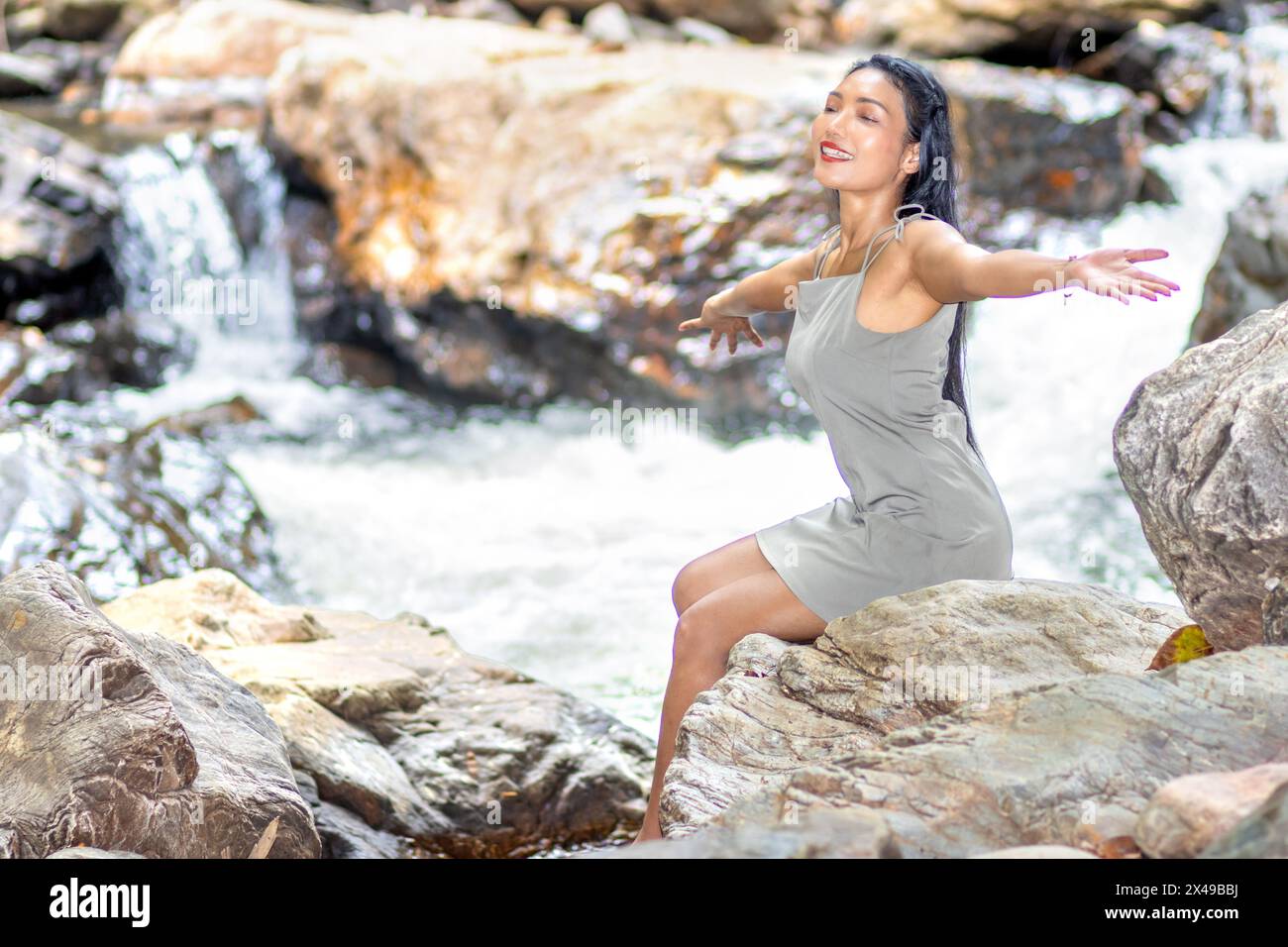 A young woman with outstretched arms sits on boulders in a water ...