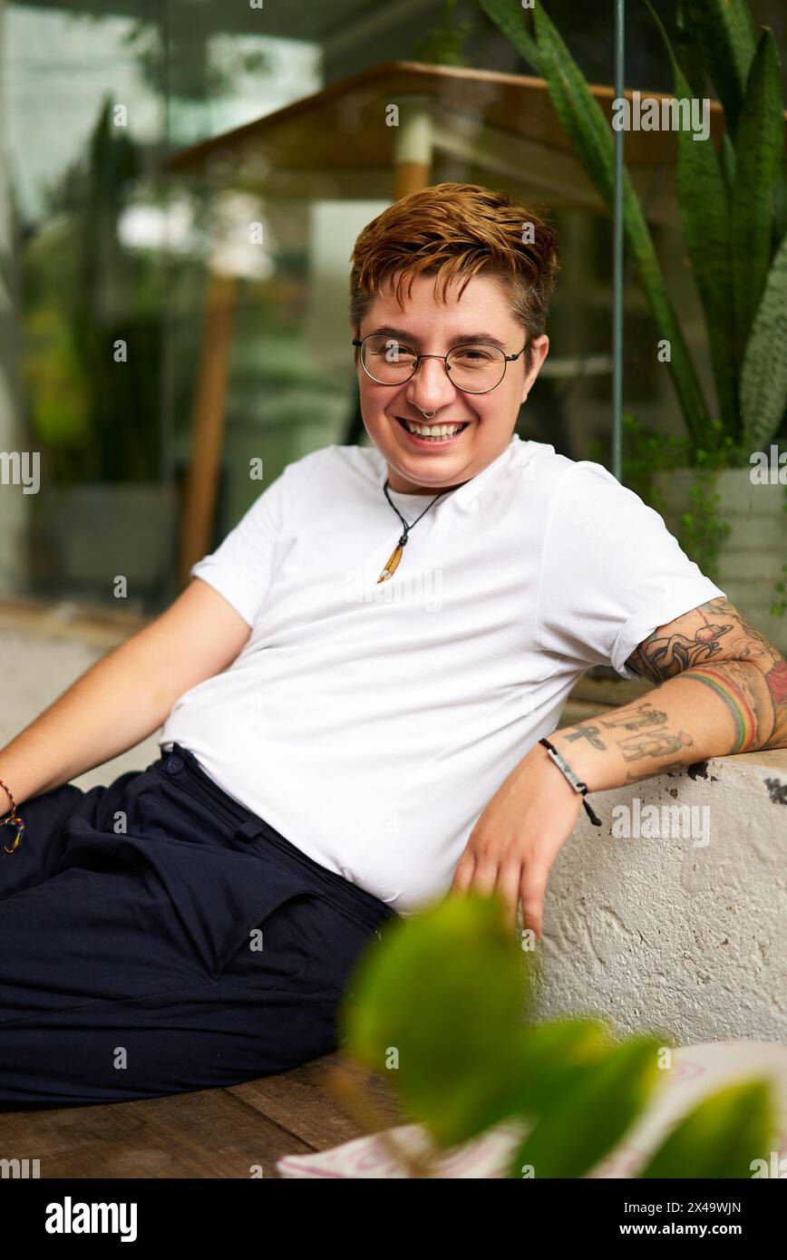 Smiling transgender person sits casually, modern coworking space with ...