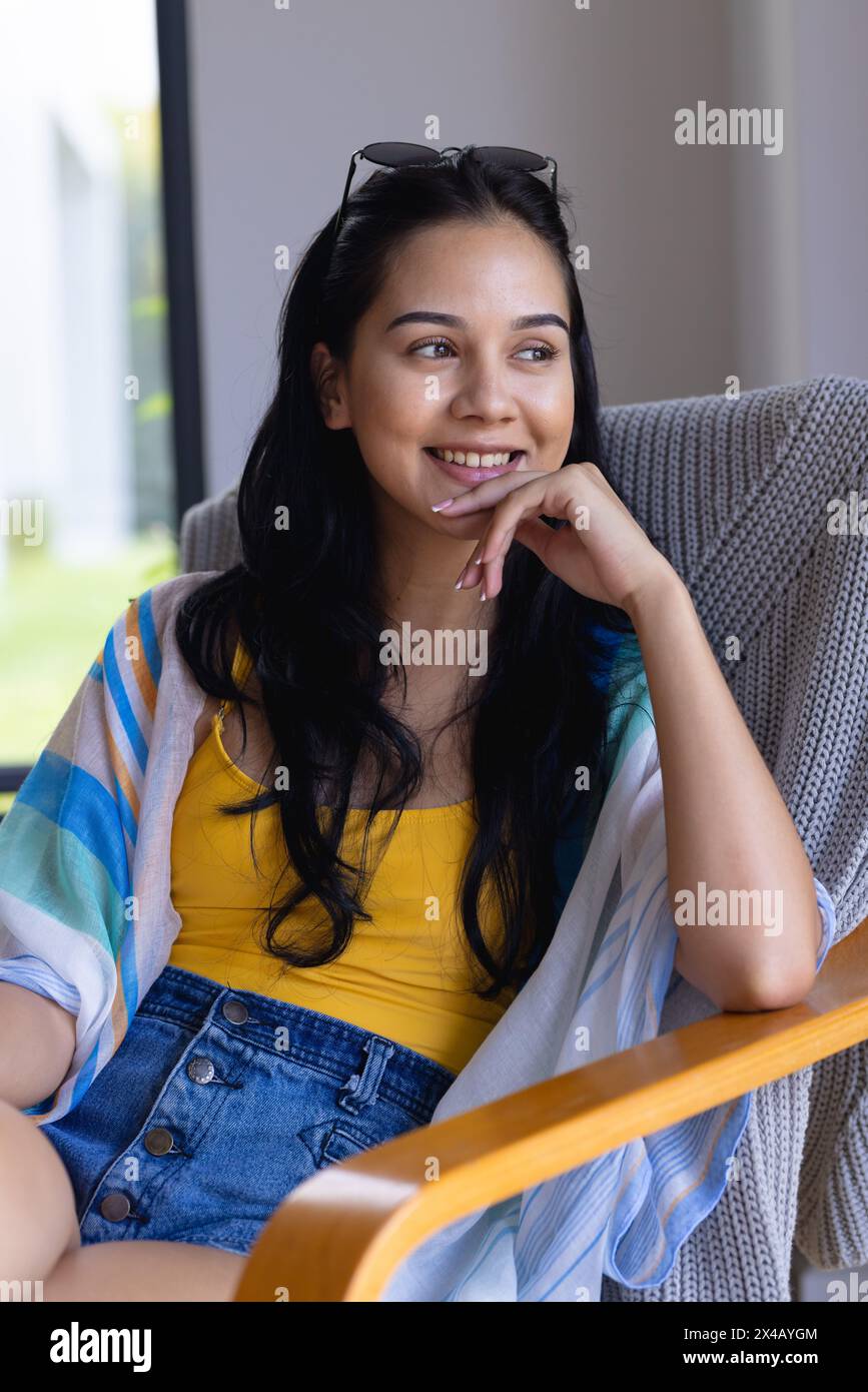 Biracial young woman sitting at home, looking away, smiling. She has long dark hair, wearing ...