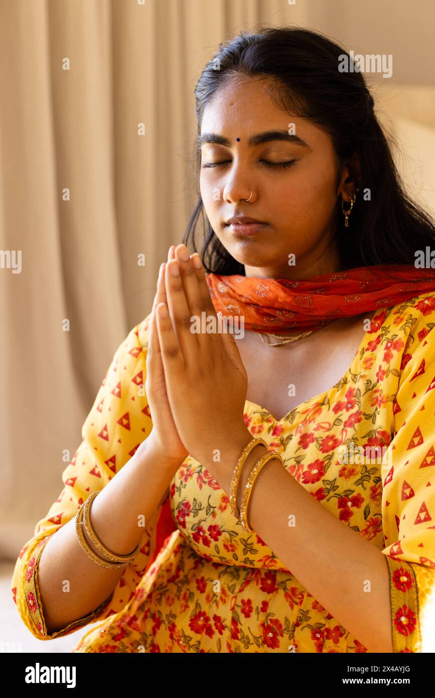 A young Indian woman, praying with eyes closed at home, wearing yellow ...