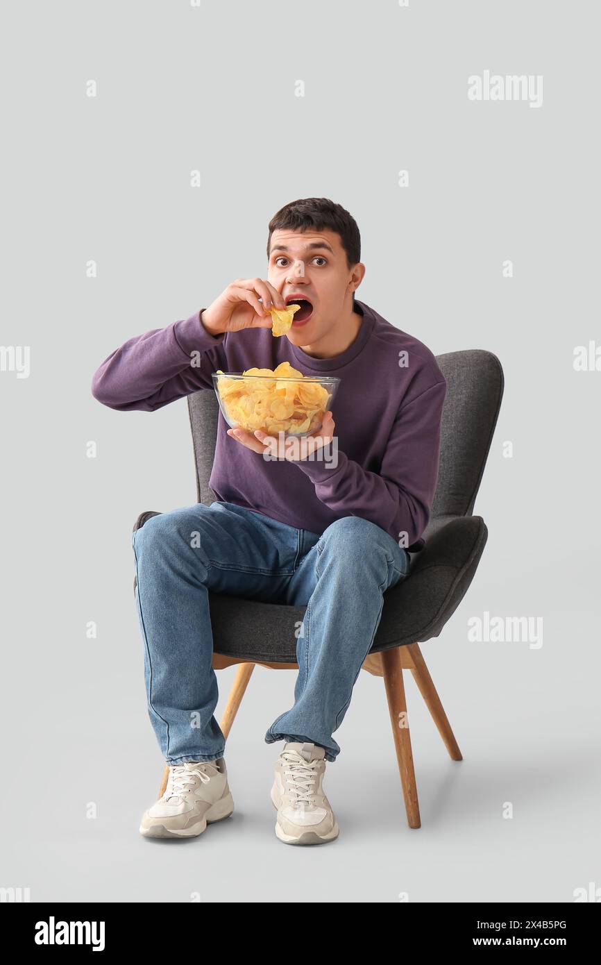 Young man eating potato chips in armchair on light background Stock ...