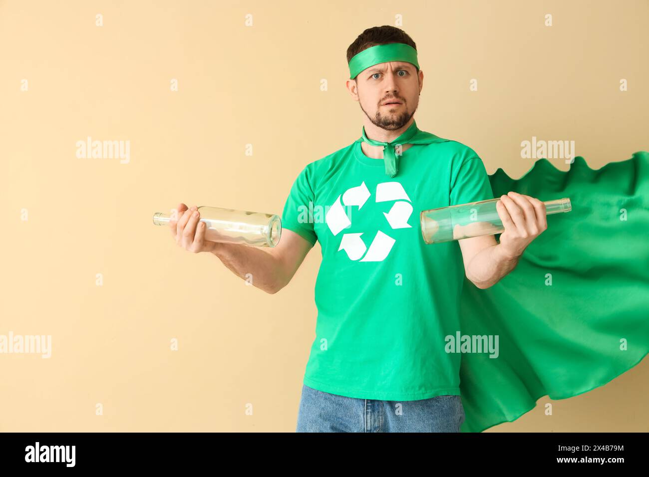 Confused young man dressed as eco superhero with glass bottles on beige ...