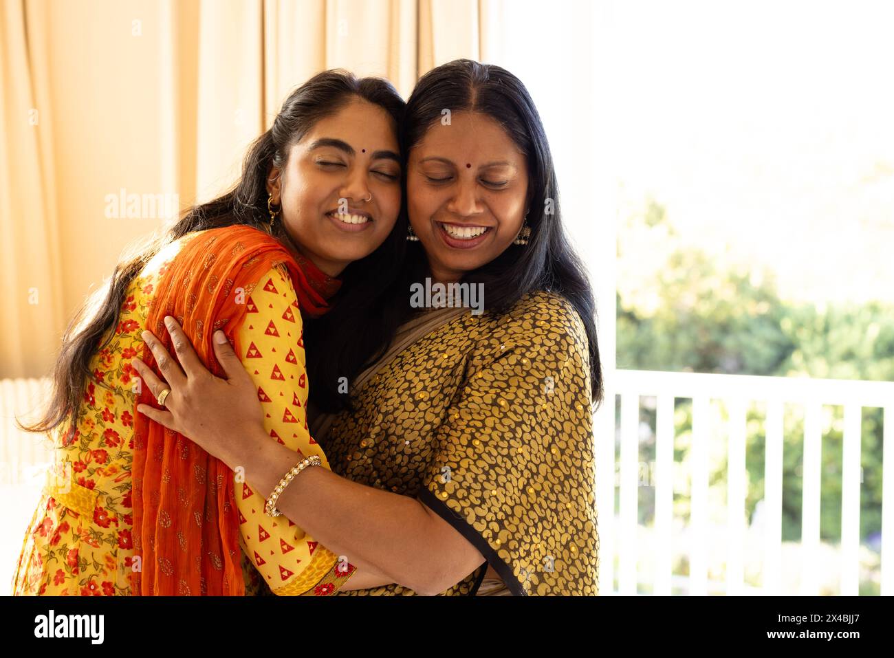 Asian mother and teenage daughter hugging at home, both smiling with eyes closed. Wearing orange ...