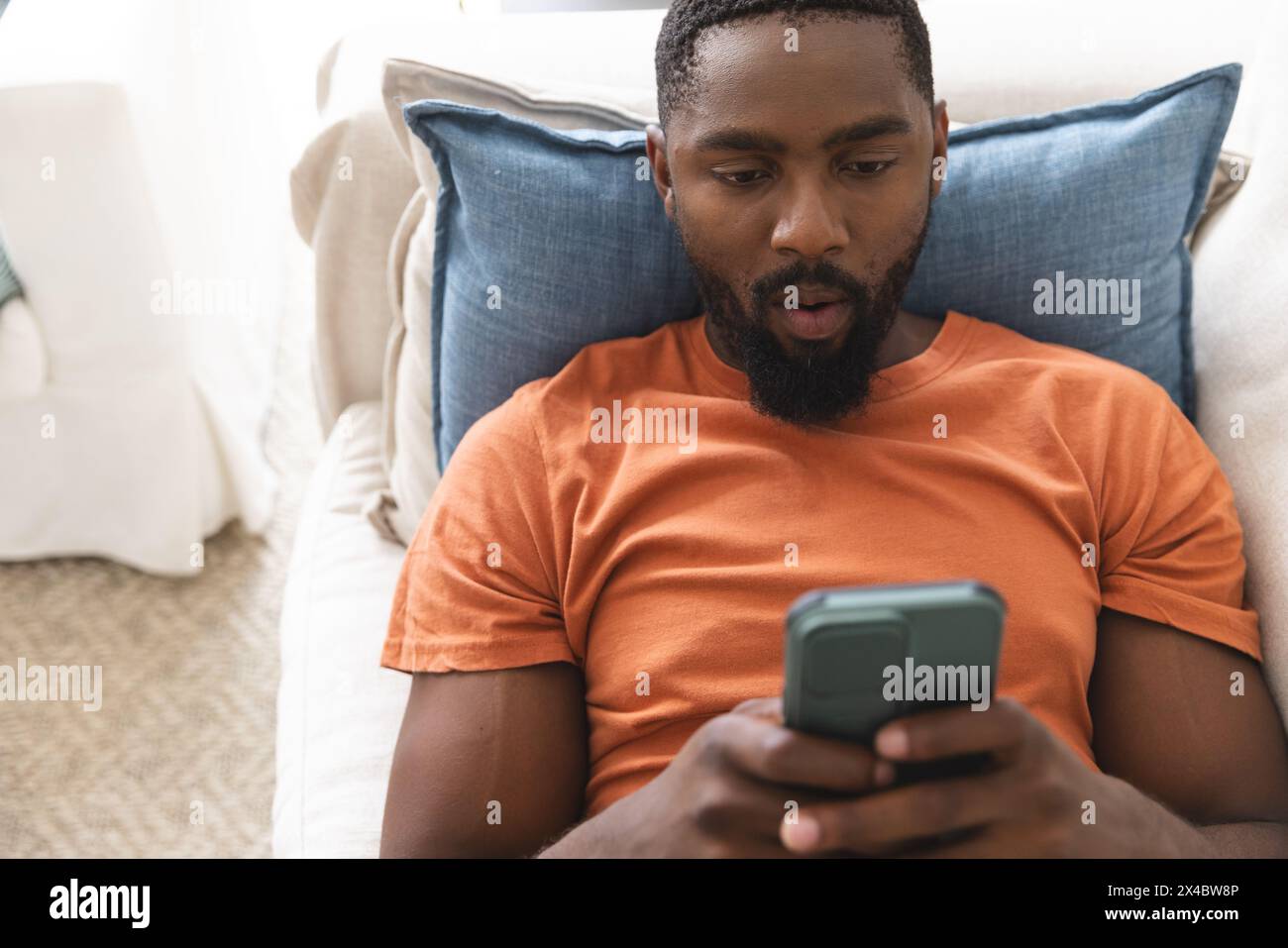 African American male homeowner lying on couch, looking at smartphone ...